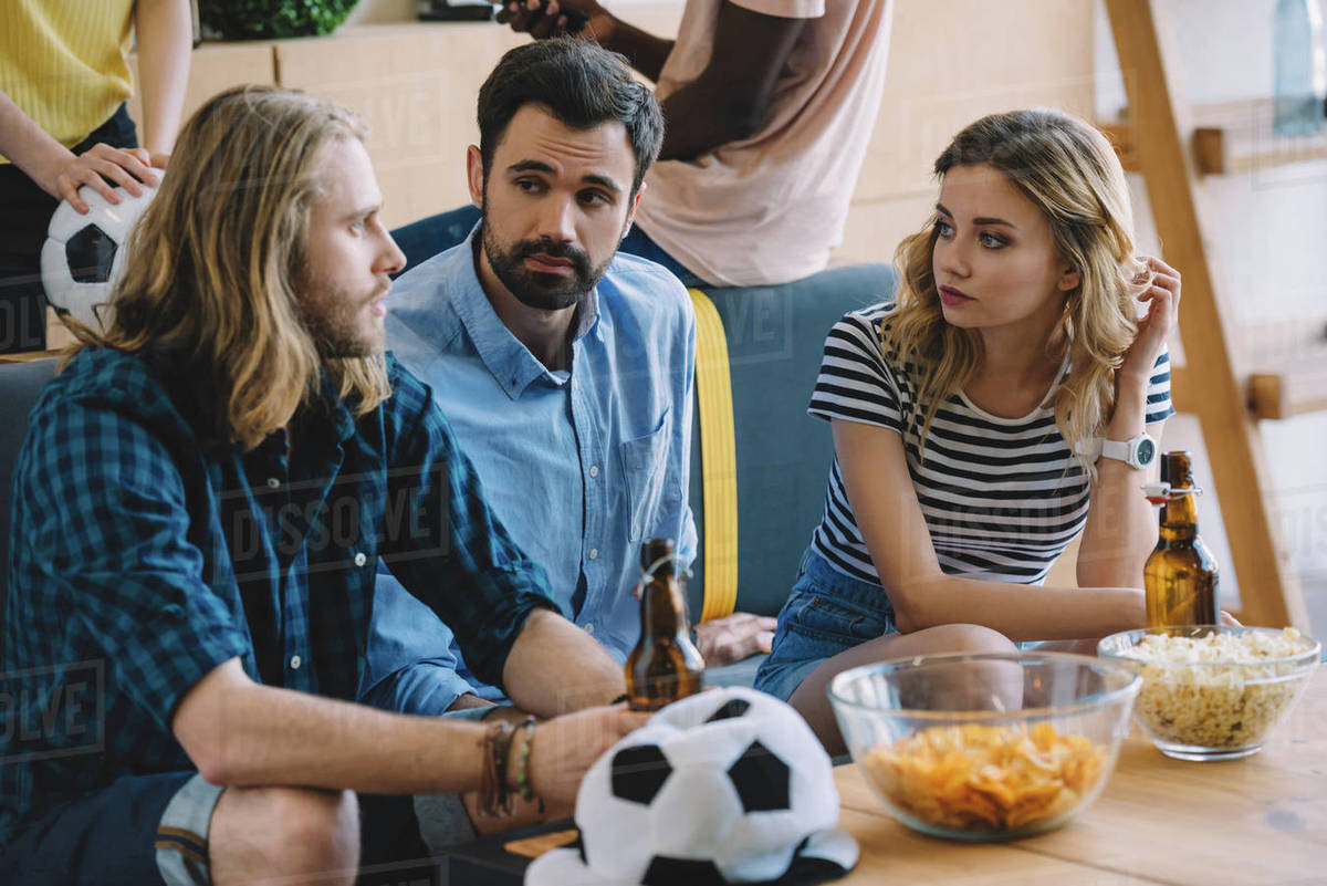 Young soccer fans sitting on sofa with beer bottles and talking to each ...