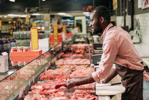 Side view of african american male butcher in apron giving steak of raw ...