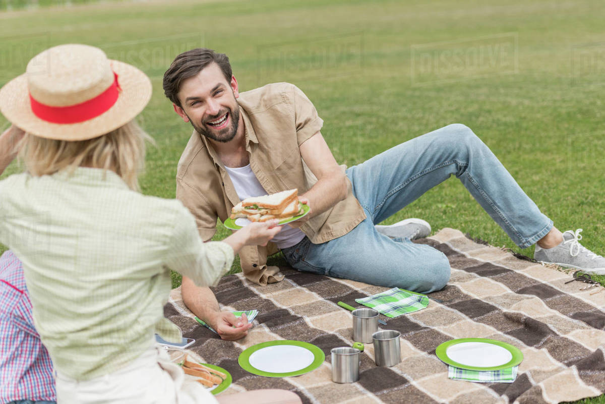Happy family eating sandwiches while sitting on plaid at picnic in park ...