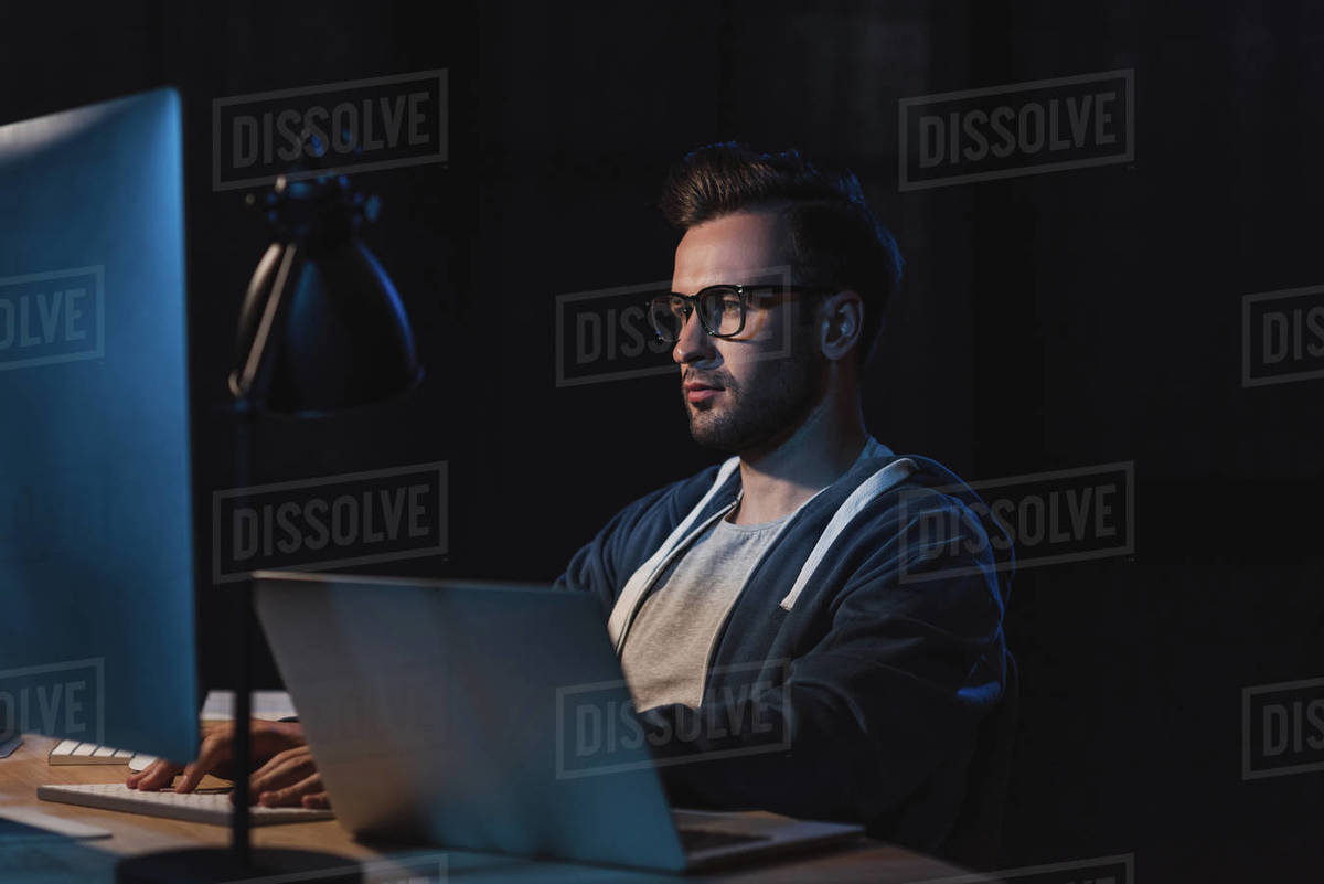 young programmer in eyeglasses working with desktop computer and laptop at night - Stock Photo ...