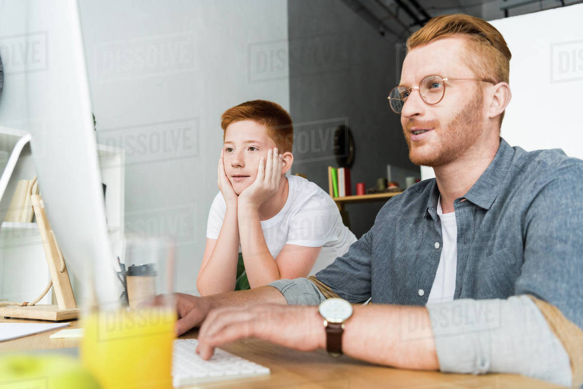 father and son sitting at table and looking at computer at home ...