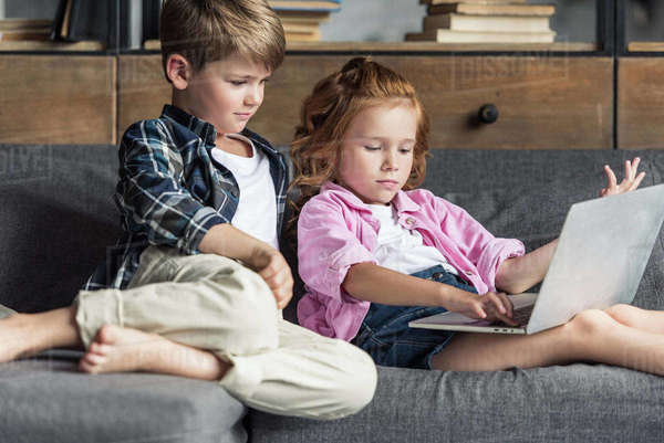 little brother and sister using laptop together while relaxing on couch ...