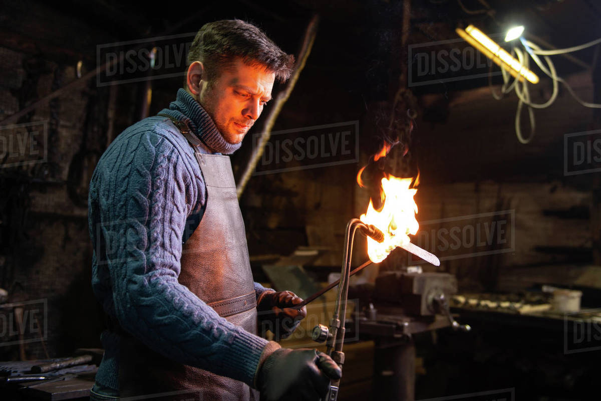 Blacksmith heating metal at flame in workshop - Stock Photo - Dissolve