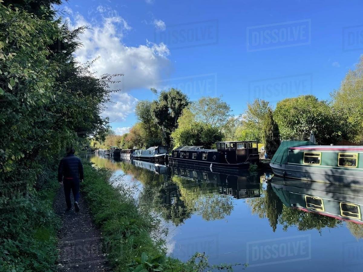 Rear view of a man walking along a towpath and Narrowboats moored along ...