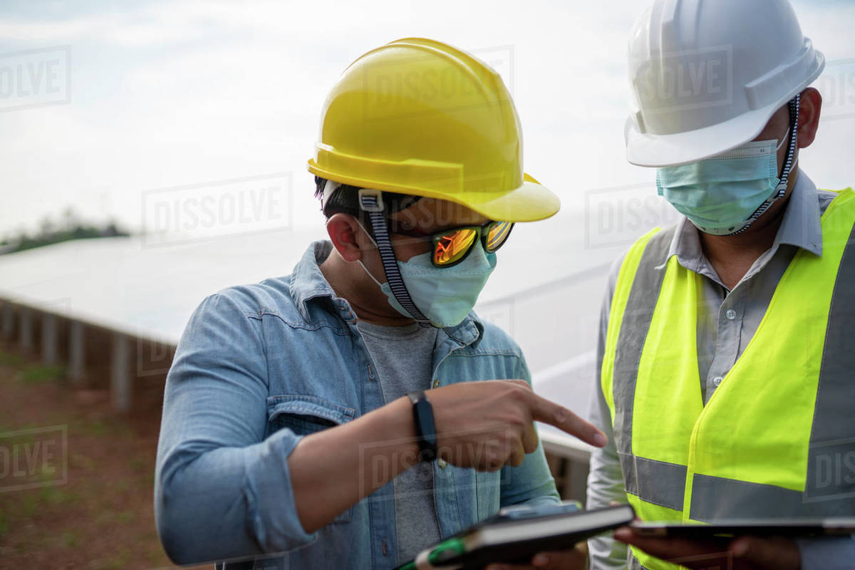 Two engineers wearing face masks looking at a digital tablet at a solar ...