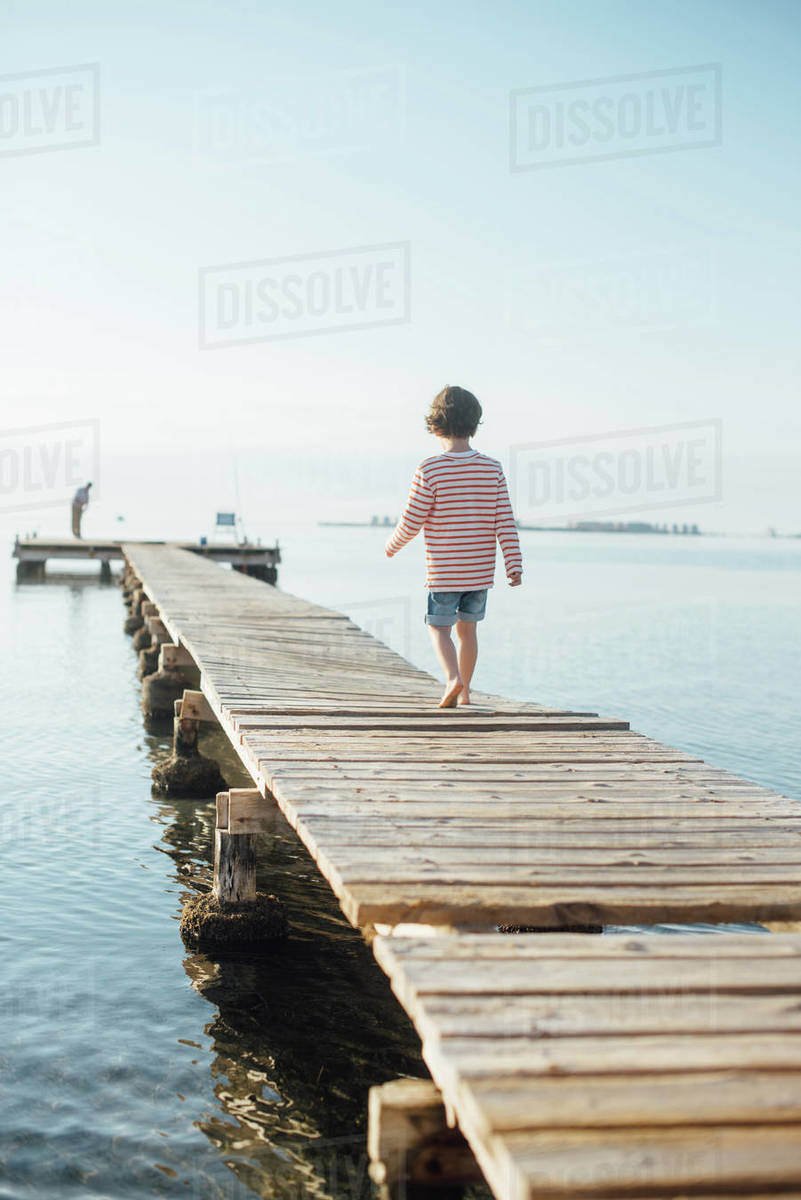 Back view of little boy walking on long wooden pier looking dreamily ...