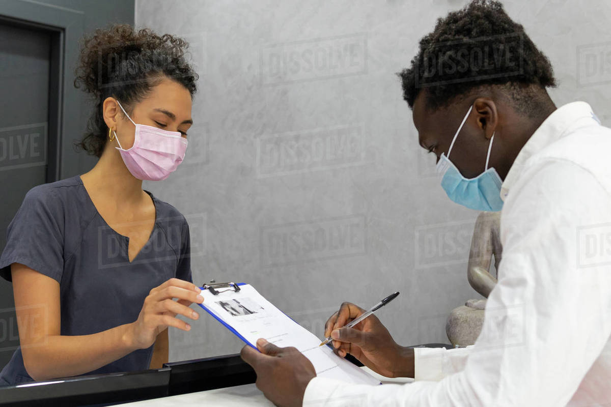 Black male patient in mask standing at reception desk near female ...