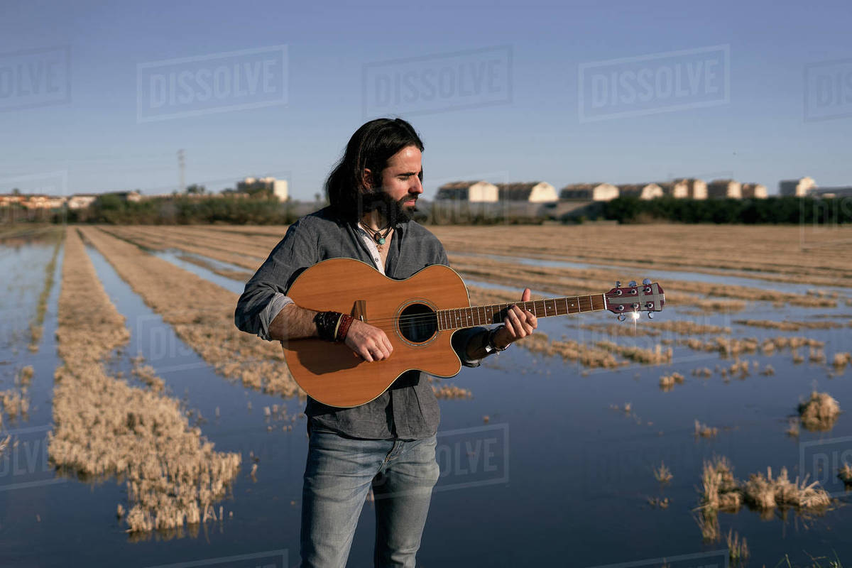 Long haired musician with beard standing on lake shore with guitar and ...