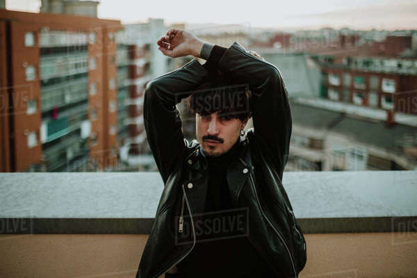 Serious man in leather jacket standing on rooftop and leaning on ...