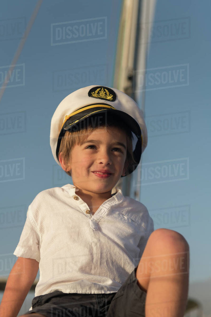 Positive kid in captain hat sitting on deck of expensive boat floating ...