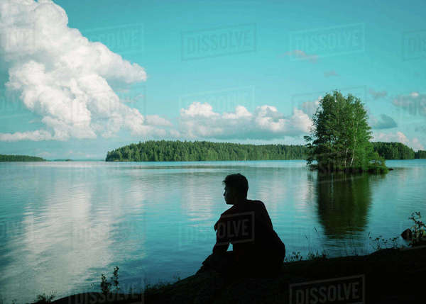 Back view of young man sitting in shadow on riverside and enjoying ...