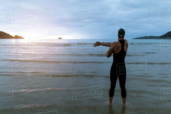 Back view of fit male swimmer in swimsuit and cap standing on seashore ...