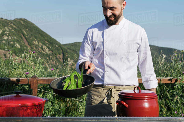 Happy bearded male cook in tunic spicing dish in pan while preparing ...