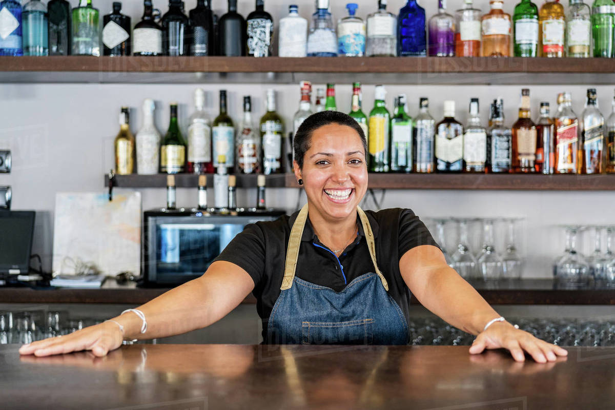 Positive female barkeeper in apron standing at counter smiling at ...