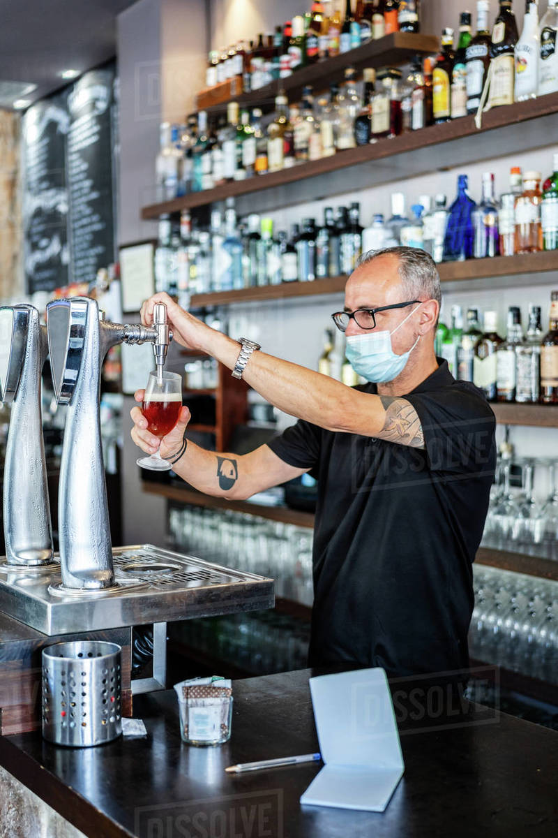 Senior ethnic male barkeeper in medical mask pouring alcohol into glass ...