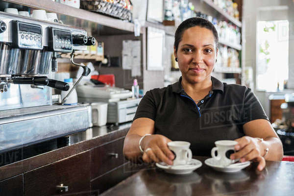 Smiling ethnic female worker in black uniform standing at bar with cups ...