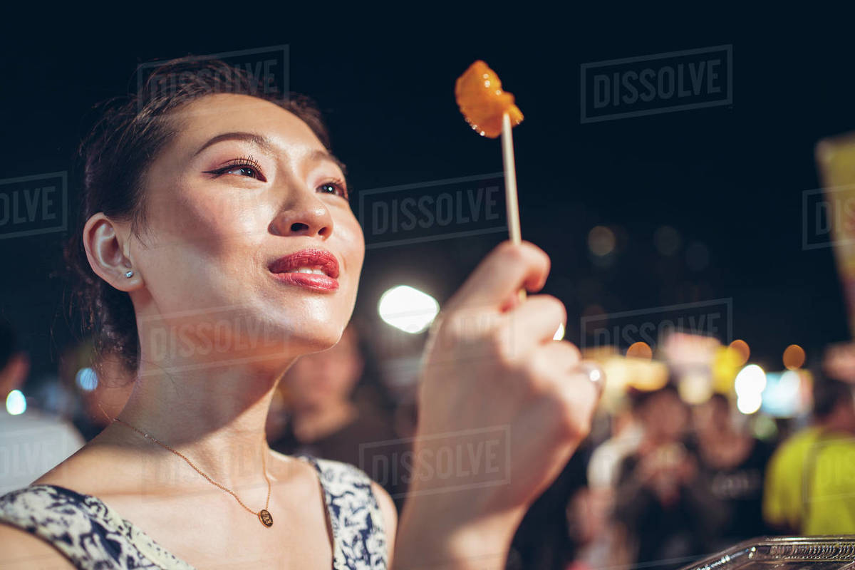 Delighted Asian female eating delicious sugar coated potato on stick ...