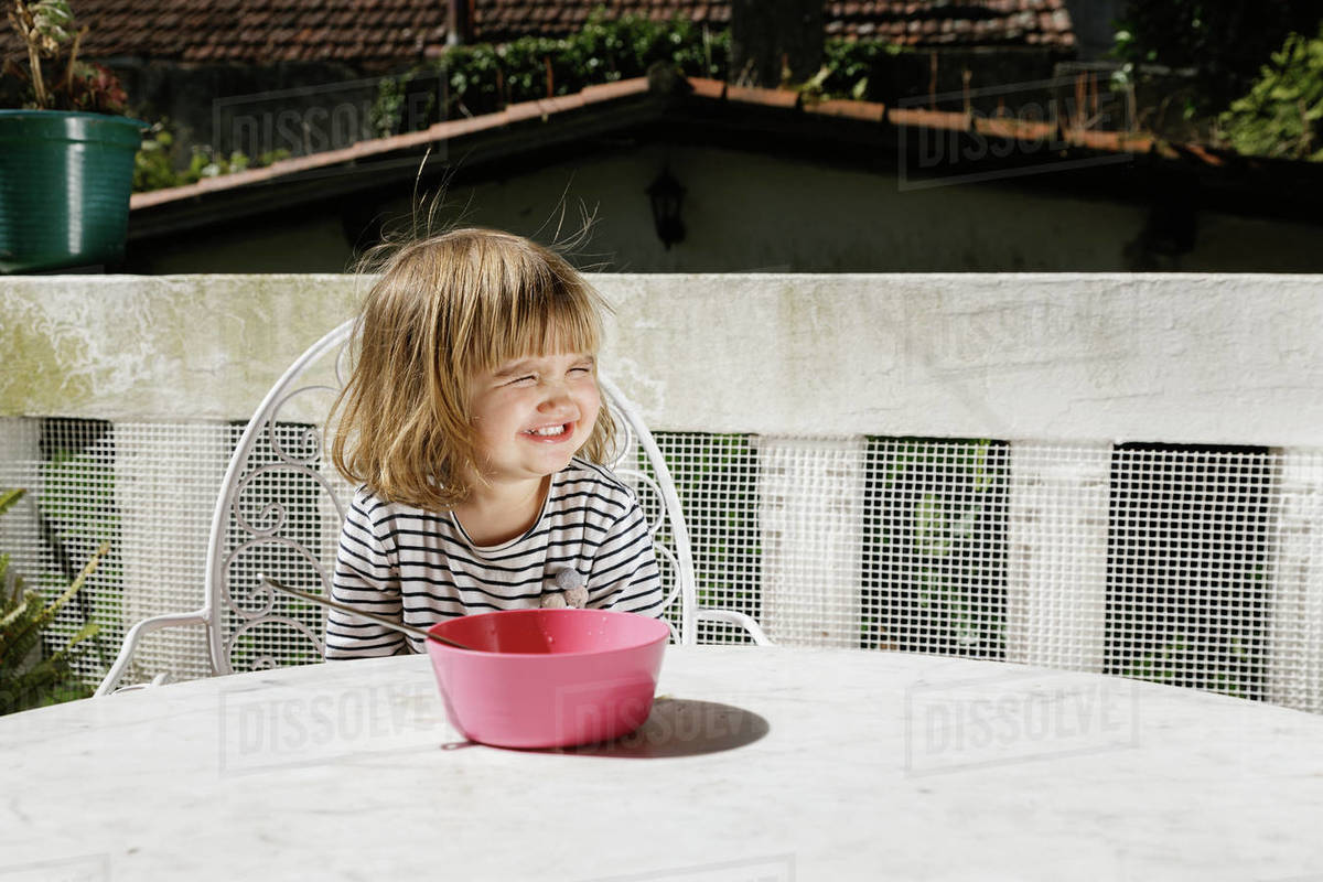 Adorable little child sitting at table with bowl on terrace and smiling ...