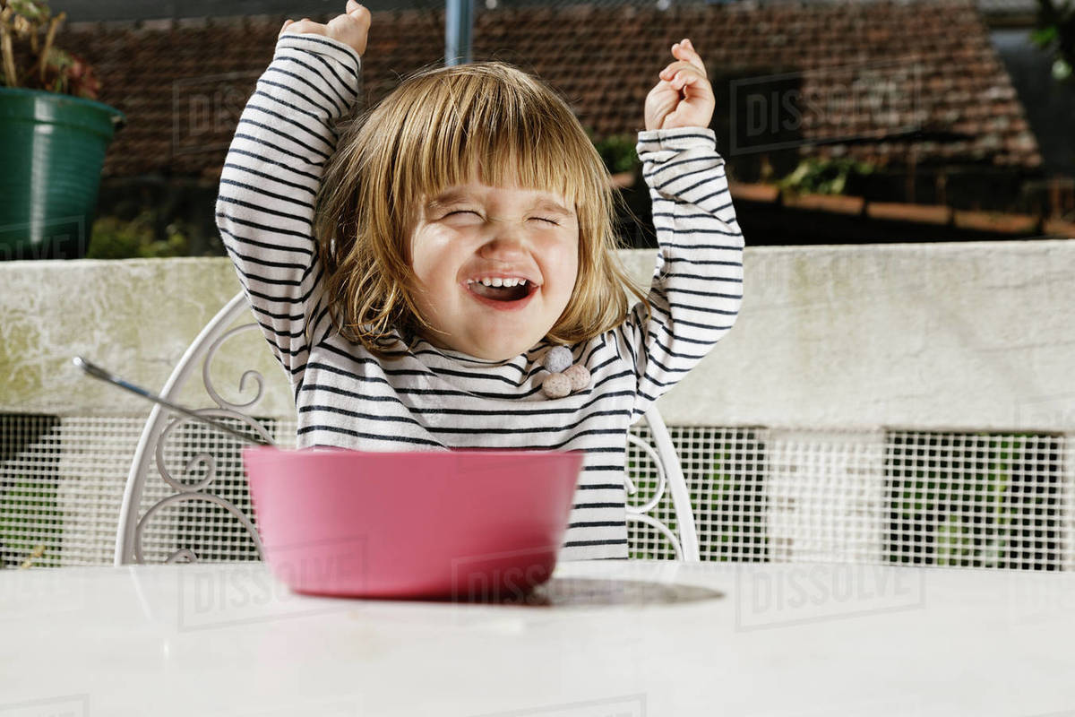 Adorable little child sitting at table with bowl on terrace and smiling ...