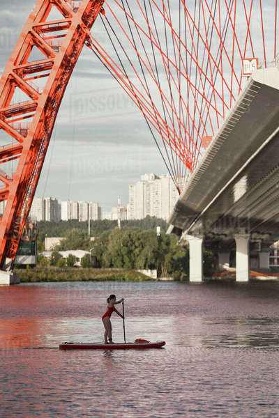 Side view of woman standing on paddle board in river and rowing with ...