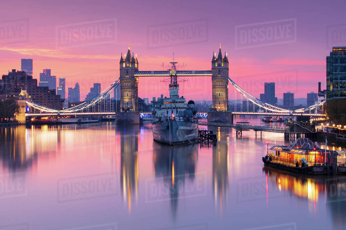 Sunrise view of HMS Belfast and Tower Bridge reflected in River Thames, with Canary Wharf in ...