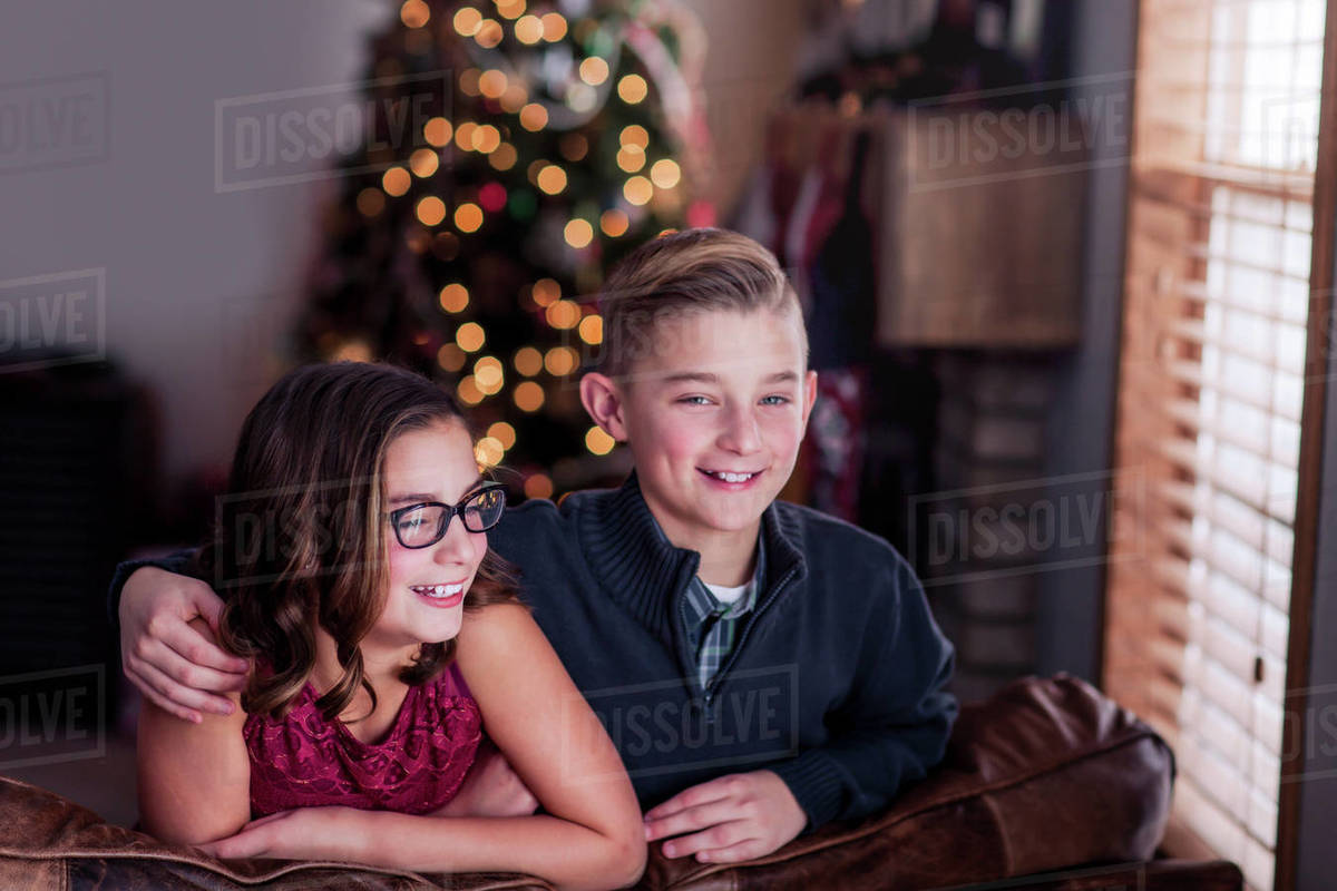 Siblings leaning over sofa, smiling, Christmas tree in background ...