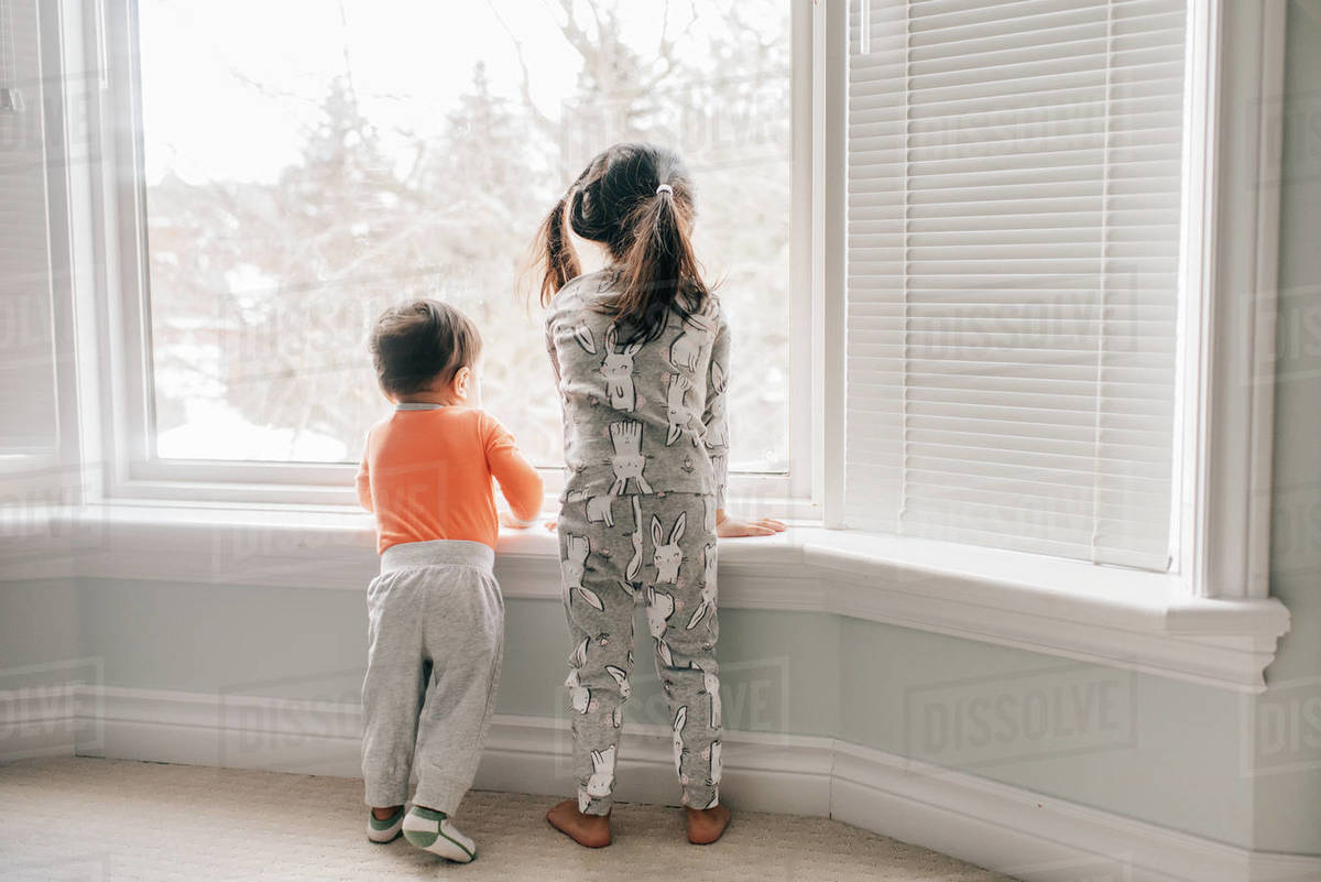 Girl and baby brother looking through living room window, rear view ...