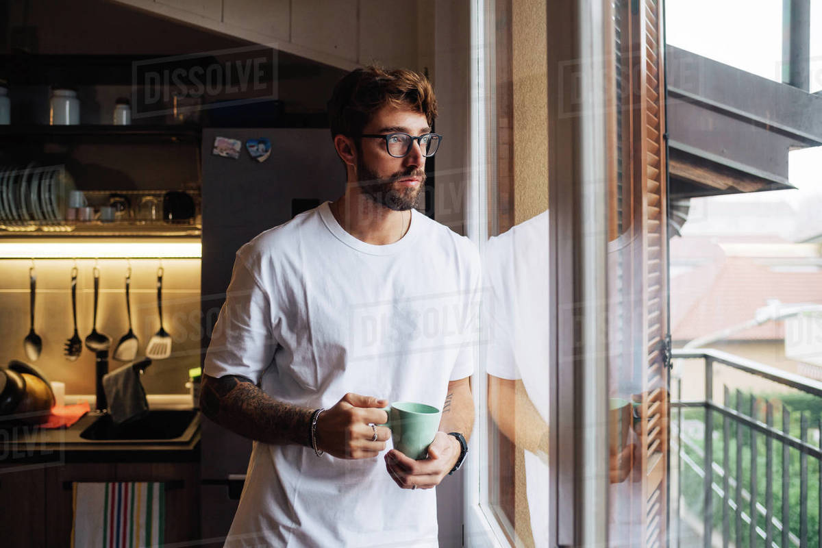 Mid adult man with coffee cup gazing out through apartment window ...