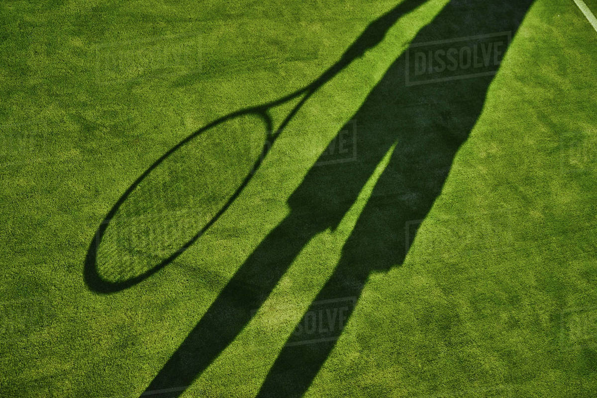 Cropped shadow of tennis player with tennis racket on green lawn ...
