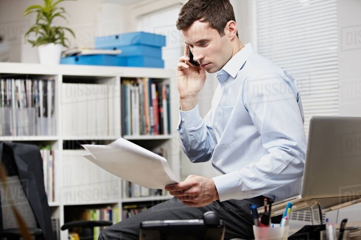 Office worker sitting on desk, on cellphone and looking at paperwork ...