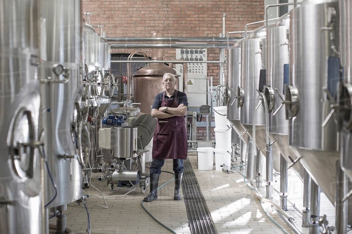 Portrait of brewer in brewery, standing next to stainless steel tanks ...