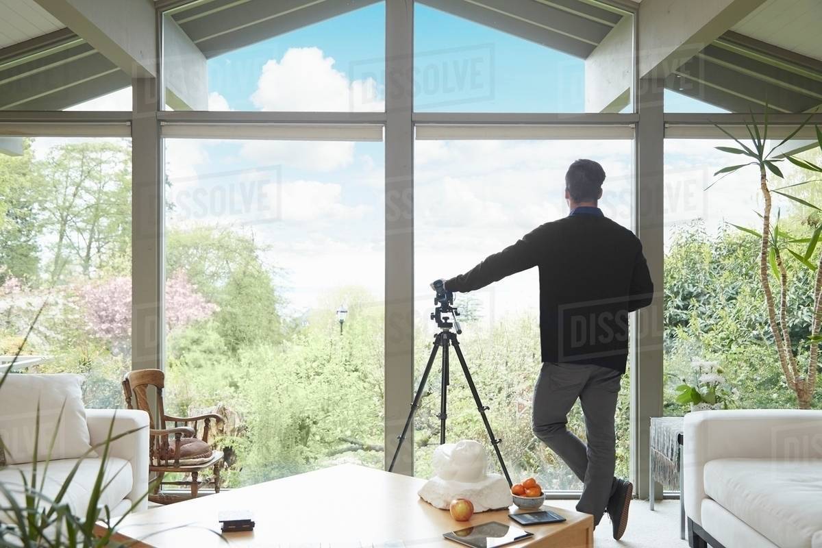 Rear view of man leaning on telescope looking out of window admiring ...