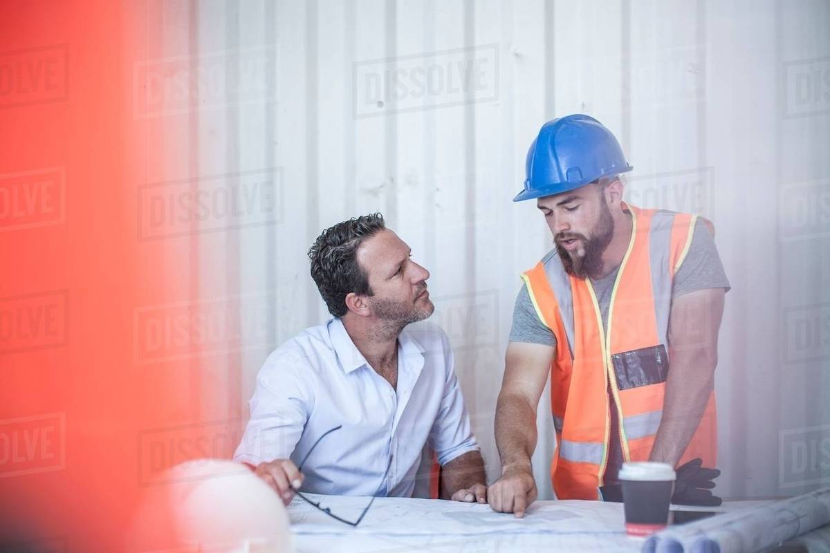 Worker explaining blueprint to construction foreman at desk in portable ...