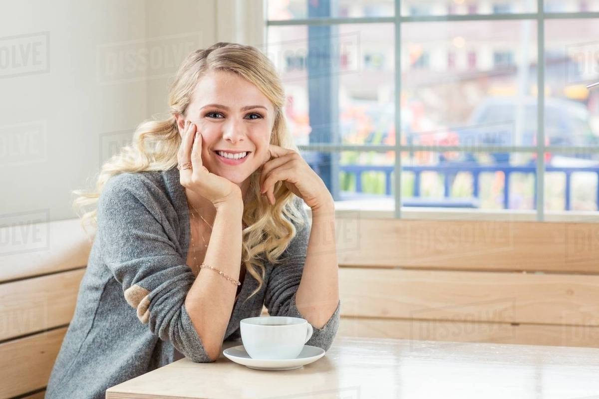 Young woman sitting at table hand on chin looking at camera smiling ...