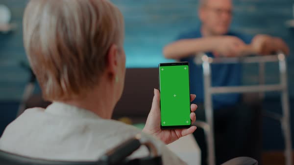 Senior woman in wheelchair holding smartphone with green screen at home ...