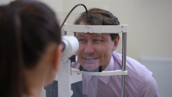 Zoom in to smiling Caucasian man sitting at slit lamp in hospital as ...