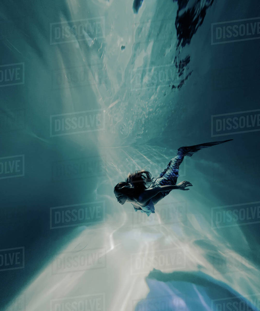 Beautiful young woman with mermaid tail swims and dives under sunlit ...