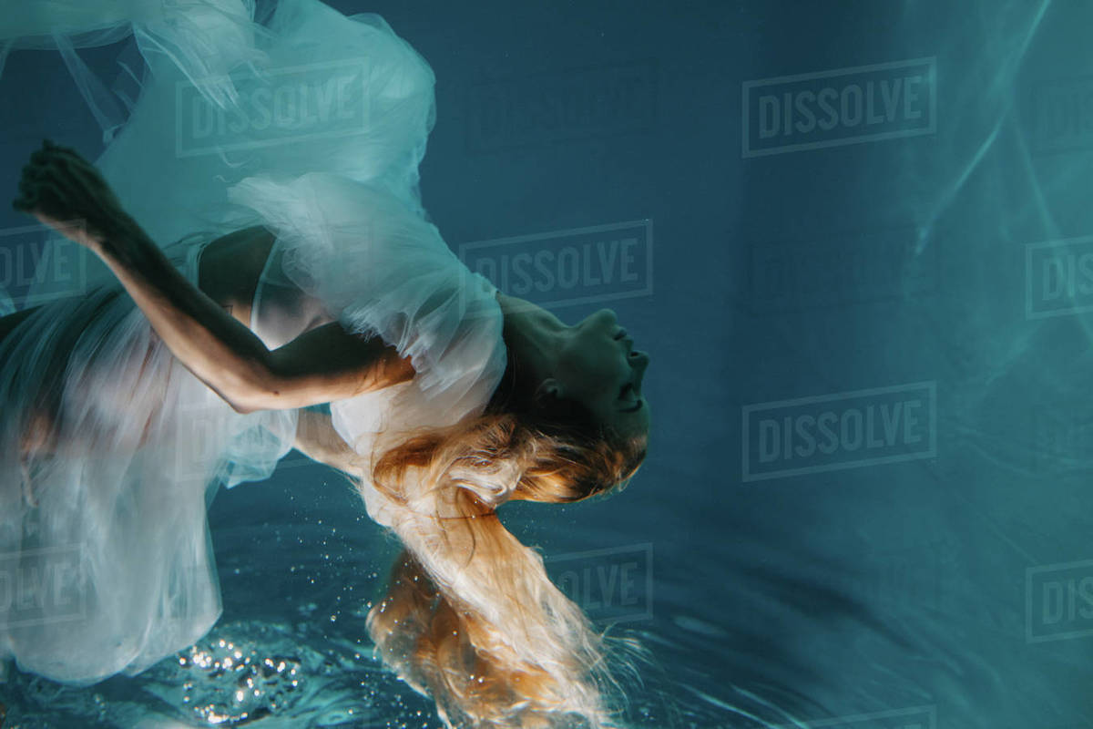Beautiful woman in bridal dress swims and dives under sunlit deep water ...