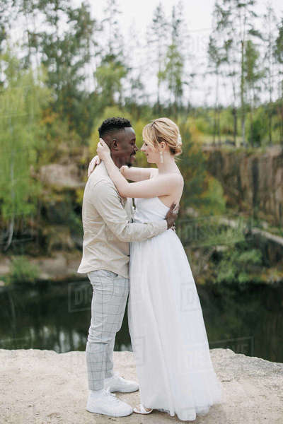 Happy interracial couple newlyweds stands on rock and embraces against ...