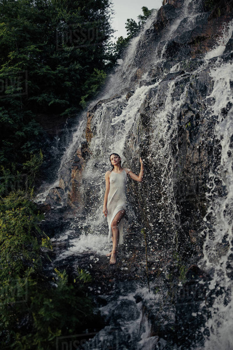 Young woman in wet dress standing near waterfall between water flows ...