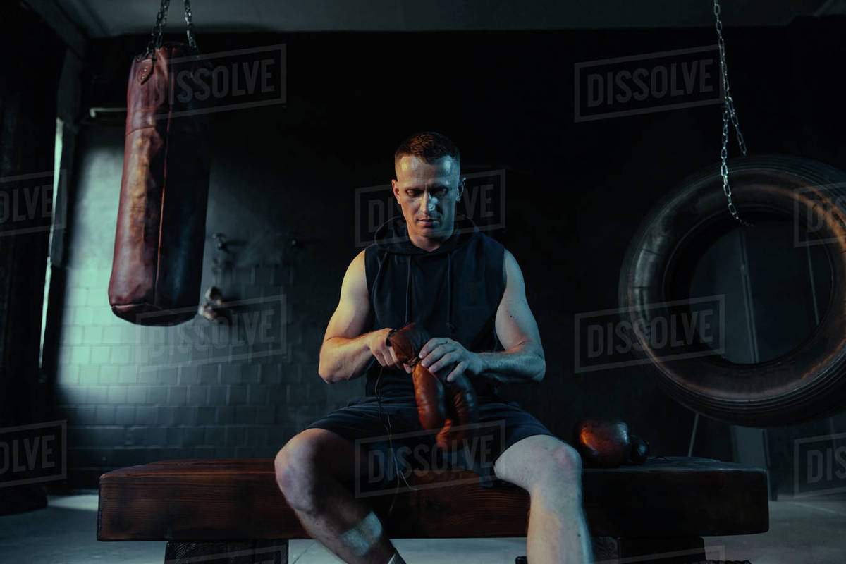 Male boxer sitting on bench in dark gym and putting on boxing gloves ...