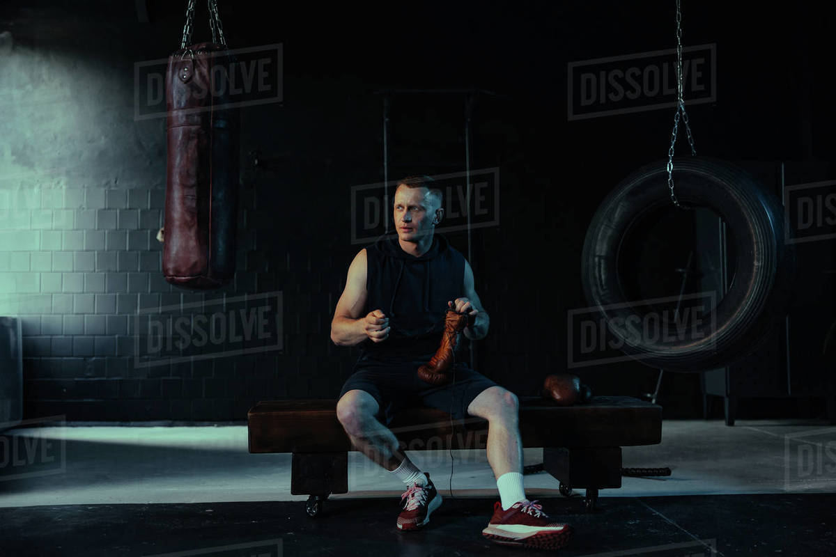 Male boxer sitting on bench in dark gym and putting on boxing gloves ...