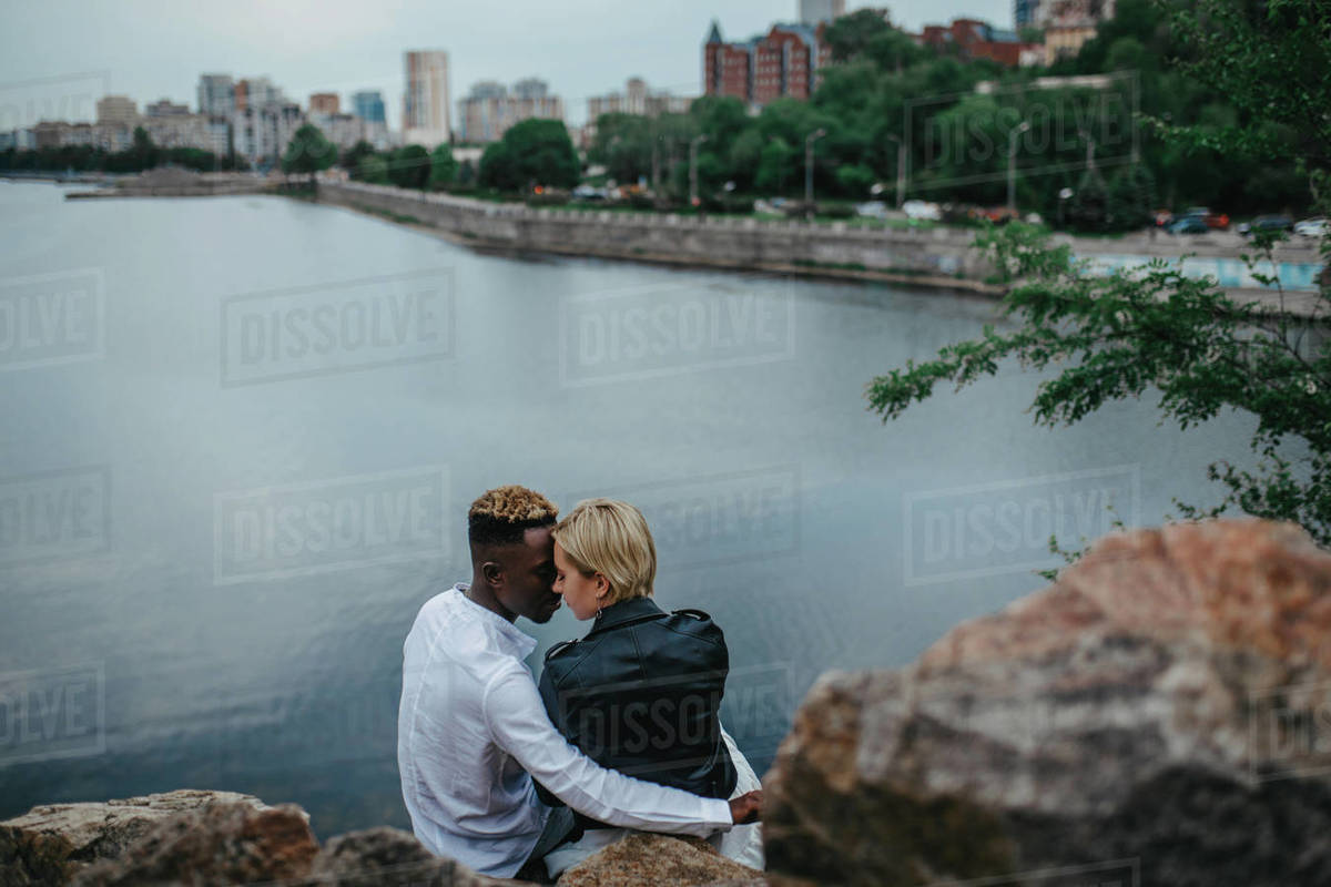 Interracial couple sits on rocks and hugs against background of river ...