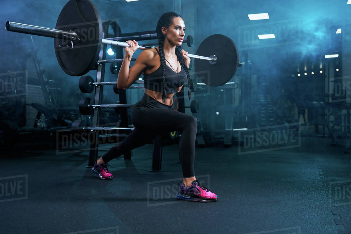 Gorgeous brunette female with long braids doing squats using barbell ...