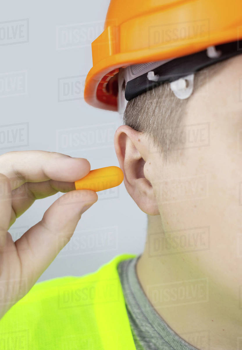 A worker in a hard hat and green vest puts on ear plugs. Industrial ...