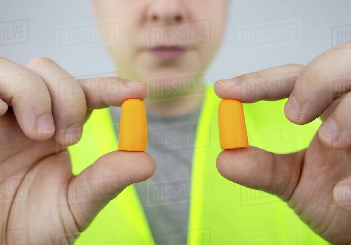 A worker in a hard hat and green vest puts on ear plugs. Industrial ...