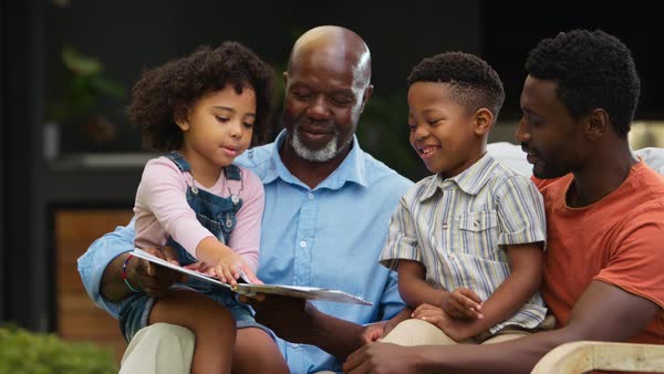 Multi-generational family sitting in garden at home having fun reading ...