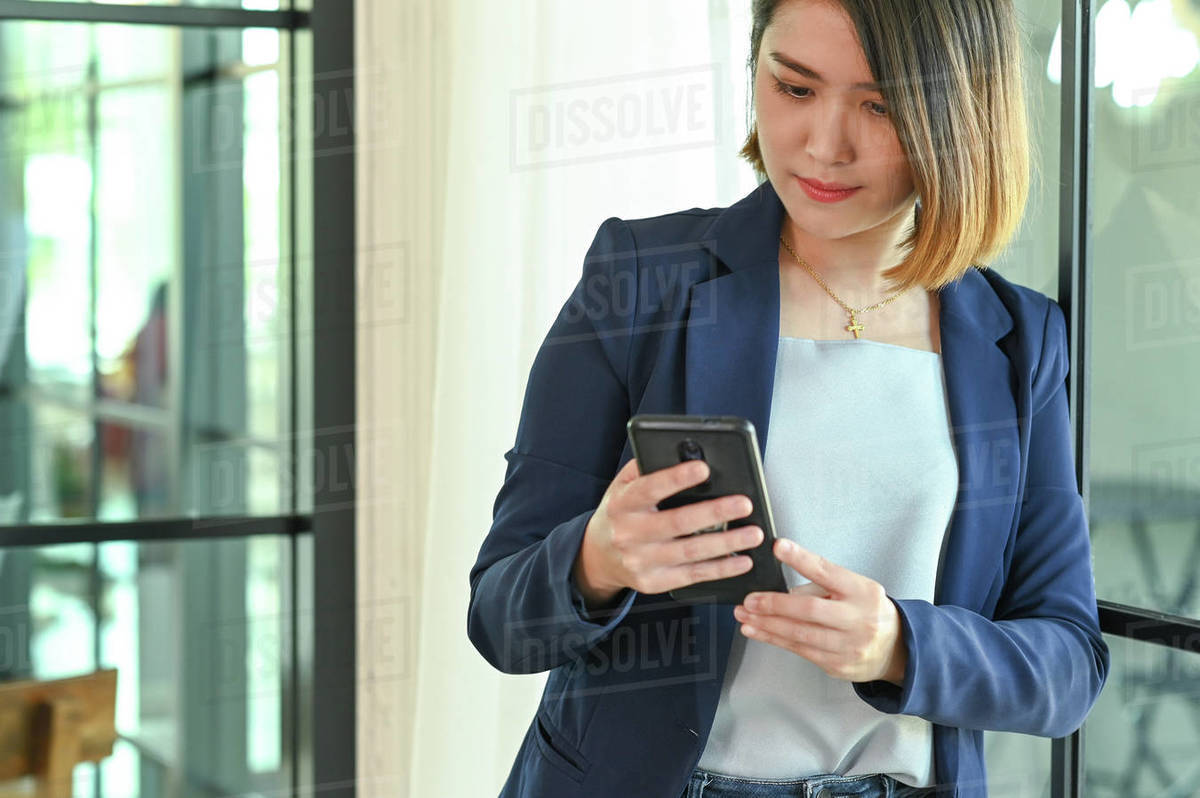 Smart woman in suit holding smartphone leaning against office window ...
