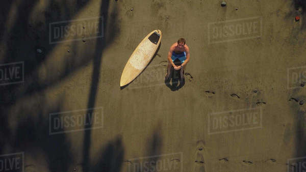 Lonely topless surfer sitting on sandy beach, looking into the horizon ...