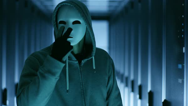 Masked hacker makes threatening gesture to the camera. He is standing ...